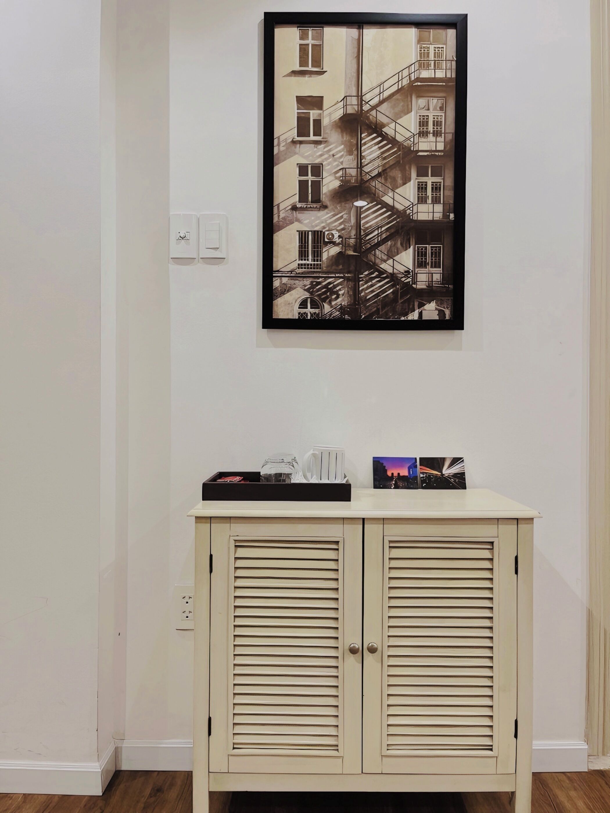 Quarto com uma obra de arte emoldurada de um edifício, uma mesinha com bandeja e um armário com portas venezianas.