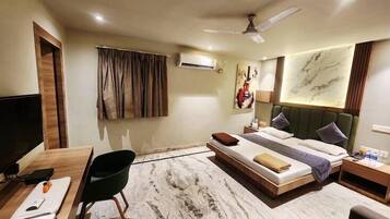Quarto de hotel com cama, escrivaninha com cadeira, televisão e ventoinha no teto.