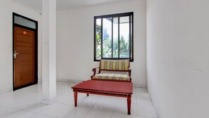 Um quarto com porta de madeira, janela com vista para vegetação, uma cadeira listrada e uma mesa vermelha.