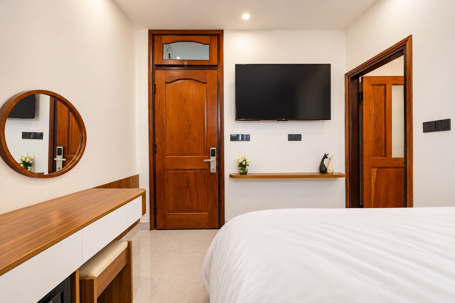 Quarto com cama, televisão fixada na parede, porta de madeira, espelho e prateleira com objetos decorativos.