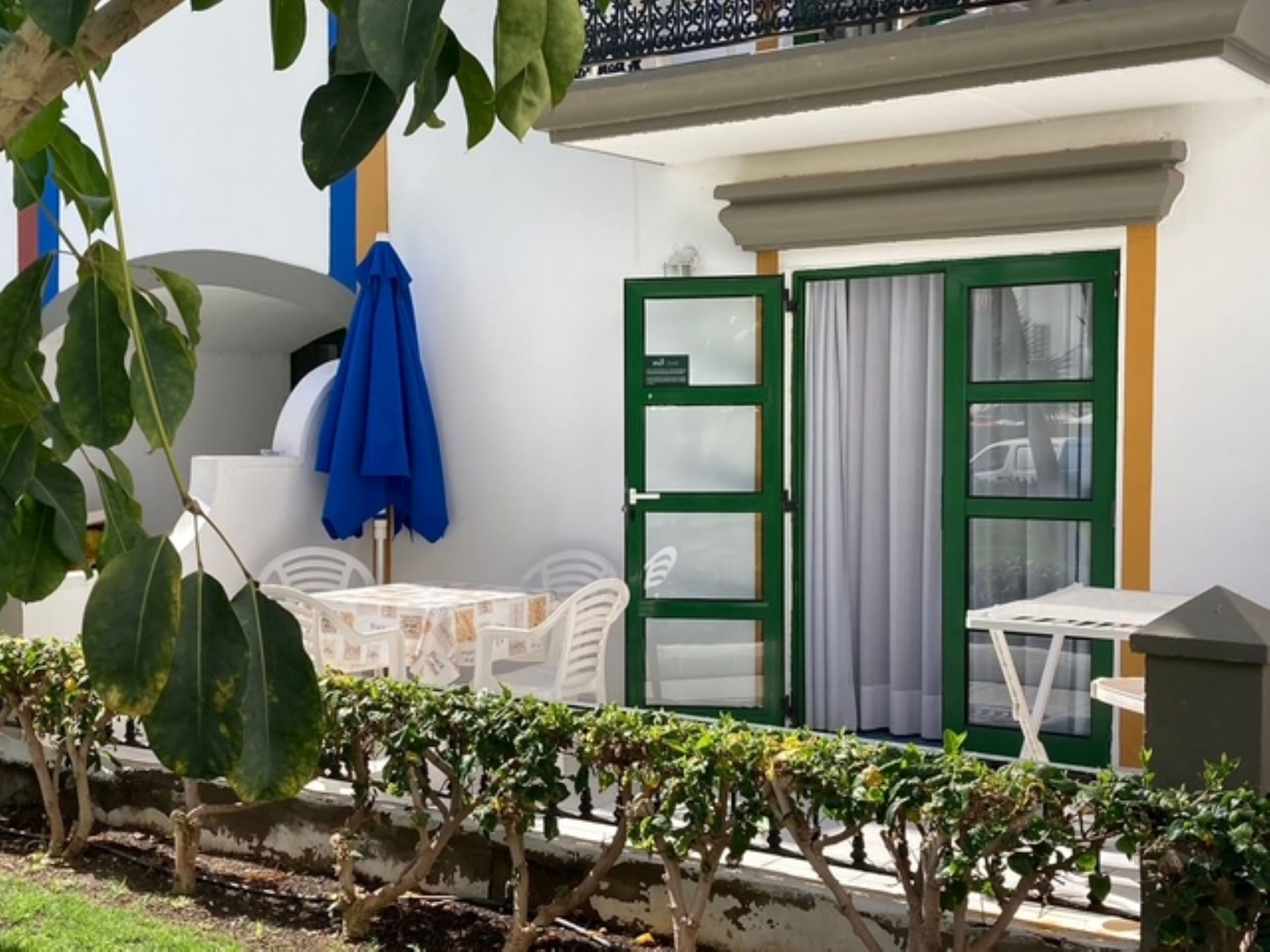 Uma pequena área externa com mesa e cadeiras, um guarda-sol azul e uma porta verde.