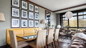 Uma sala de jantar com um banco amarelo, uma mesa de vidro e cadeiras. Há quadros nas paredes, uma janela grande com vista e um lustre.