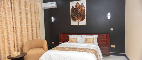Quarto de hotel com cama, uma cadeira, uma mesinha, ar condicionado e uma obra de arte na parede.