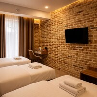 Quarto de hotel com duas camas, uma escrivaninha, uma cadeira, televisão de tela plana presa a uma parede de tijolos e uma janela com cortinas.