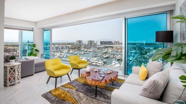 Uma sala de estar ampla com uma grande janela com vista para um porto e para o horizonte da cidade.