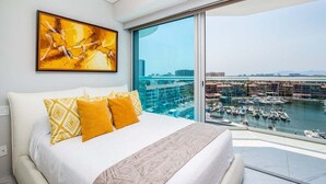 Quarto de hotel moderno com uma cama grande, vista da cidade através de uma janela do chão ao teto, e um porto com barcos à vista.