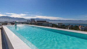 Uma piscina infinita no terraço com vista clara para o oceano e montanhas ao fundo.