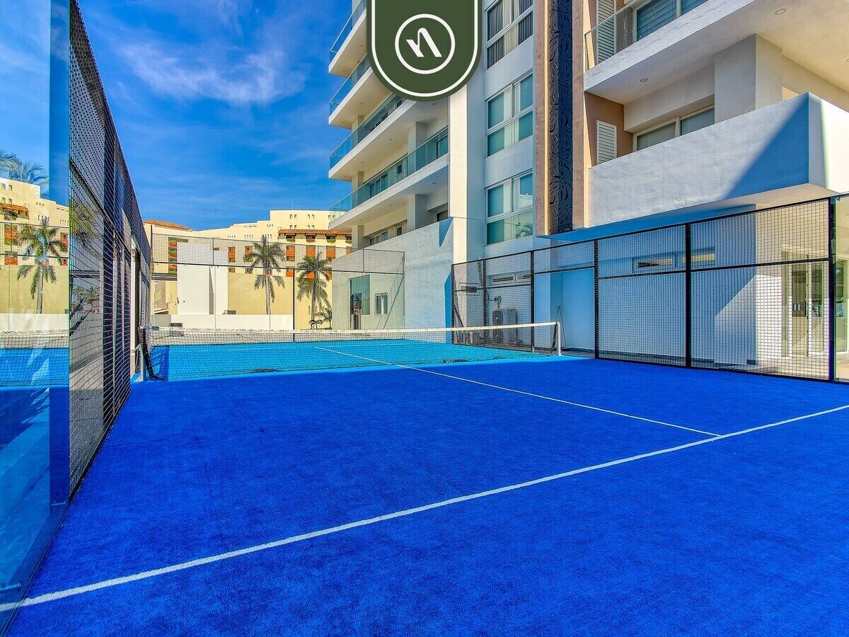 Uma quadra de tênis com rede, cercada por uma piscina e edifícios.