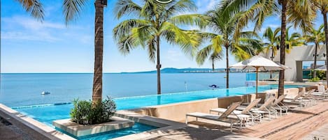 Área de piscina com espreguiçadeiras e palmeiras, com vista para um corpo de água.