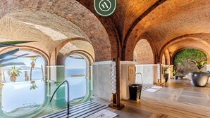 Área de piscina interior luxuosa com vista para o mar, apresentando tetos de tijolo em arco e uma grande planta em vaso.