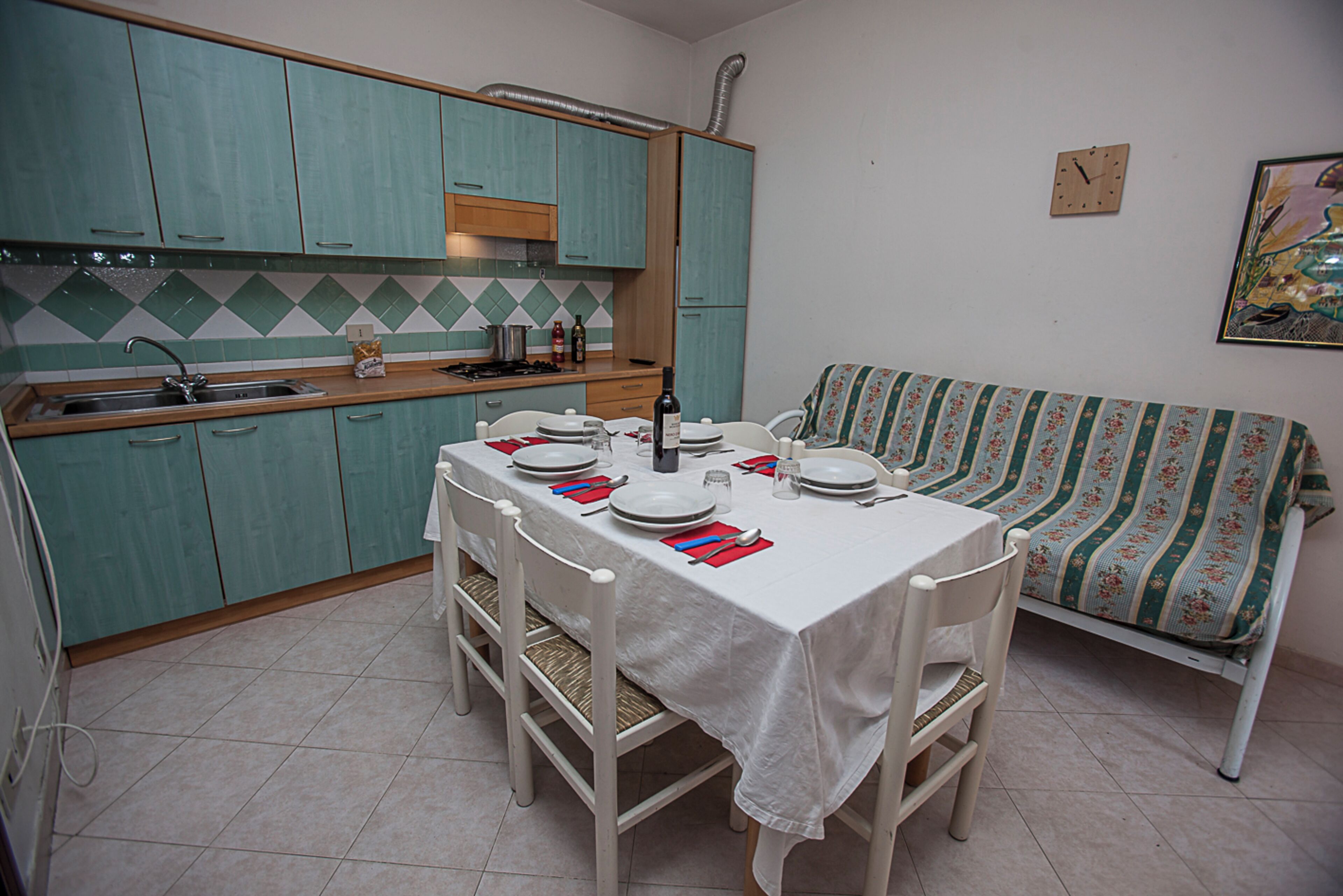 Uma cozinha compacta com armários verde-água, uma mesa de jantar posta para quatro pessoas e um sofá estampado no canto.