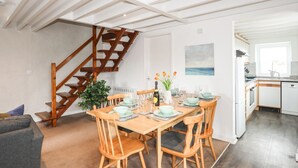 Uma sala de jantar com mesa de madeira posta para quatro pessoas, rodeada por cadeiras de madeira. Uma escada com corrimão de madeira leva a um andar superior.