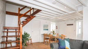 Uma sala ampla com escada de madeira, mesa de jantar para quatro pessoas e um sofá cinza com almofadas coloridas.