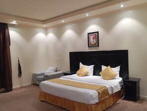 Quarto de hotel com uma cama grande, uma poltrona cinza, uma mesa pequena e um quadro na parede.