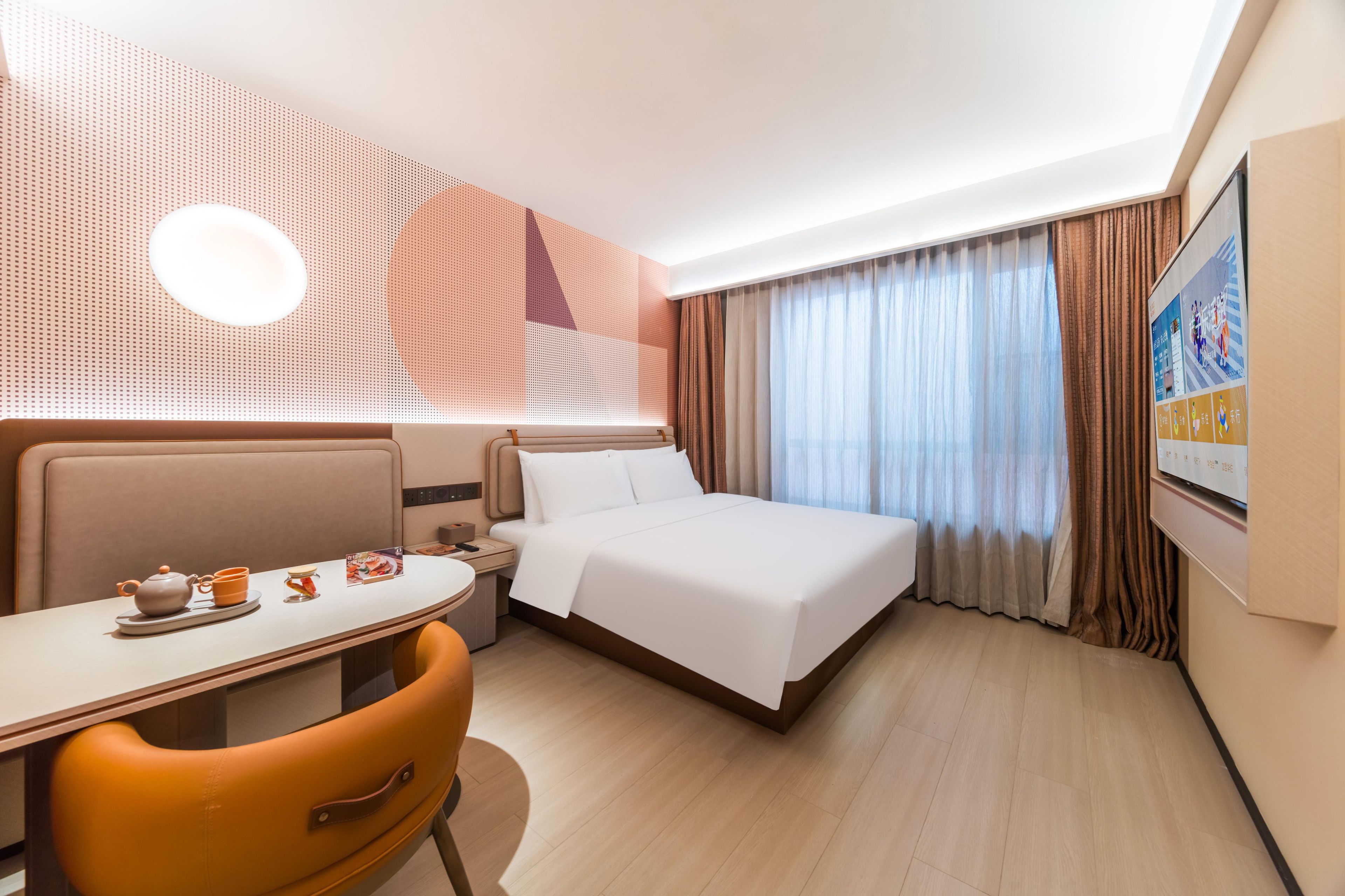 Quarto de hotel com uma cama, uma mesa com cadeira, uma mesinha com bule e chávenas, e uma televisão na parede.