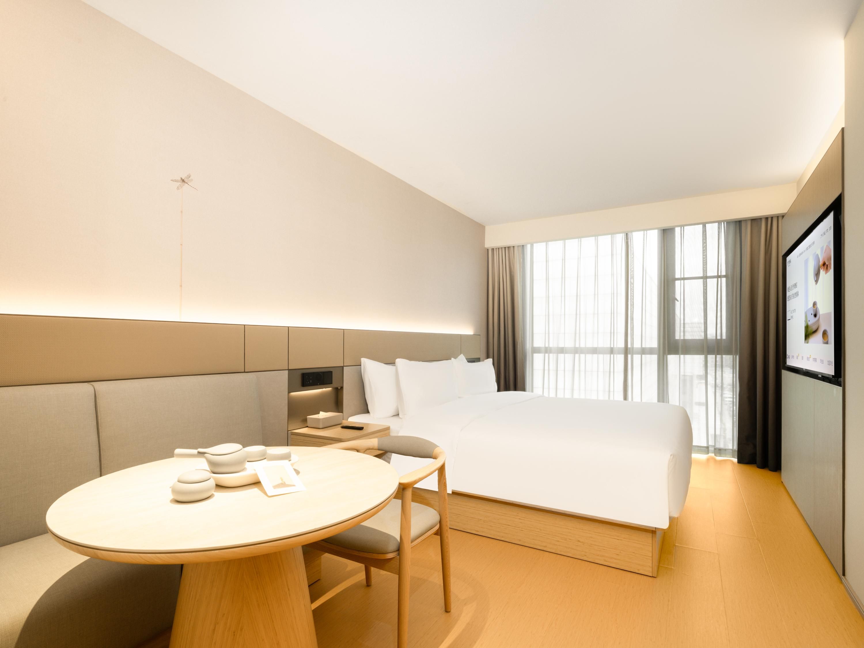 Quarto de hotel moderno com uma cama grande, uma mesa de cabeceira, uma cadeira, uma mesa redonda com um controle remoto e uma TV na parede.