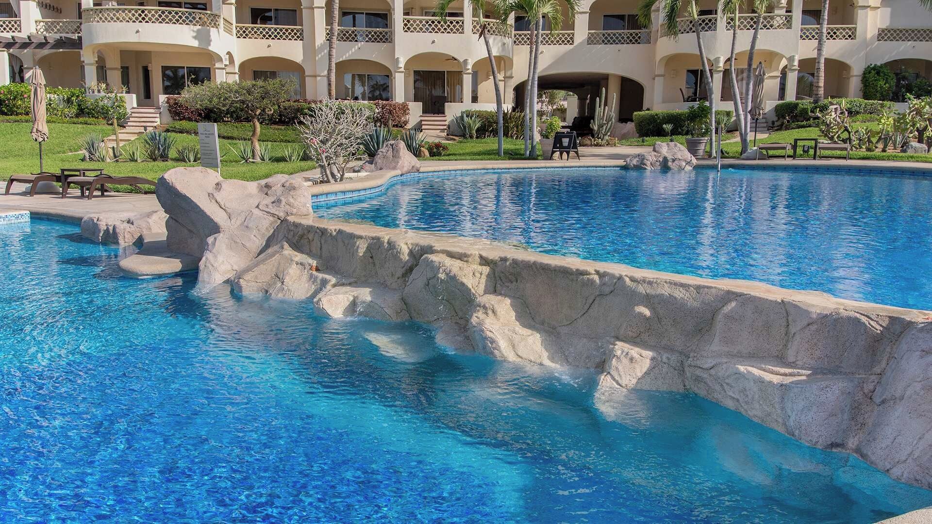 Piscina com borda rochosa, rodeada por um edifício com várias varandas e um jardim bem cuidado.