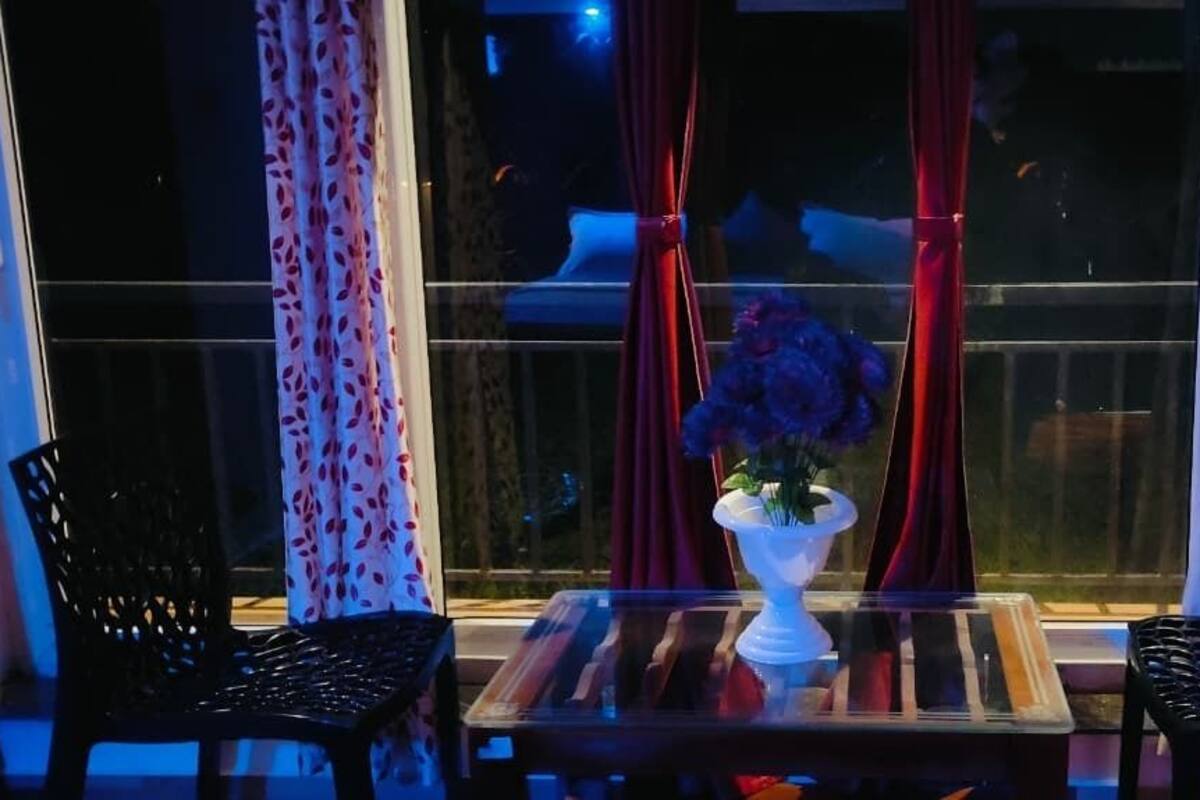 Uma mesa com um vaso de flores, duas cadeiras e vista pela janela com cortinas.