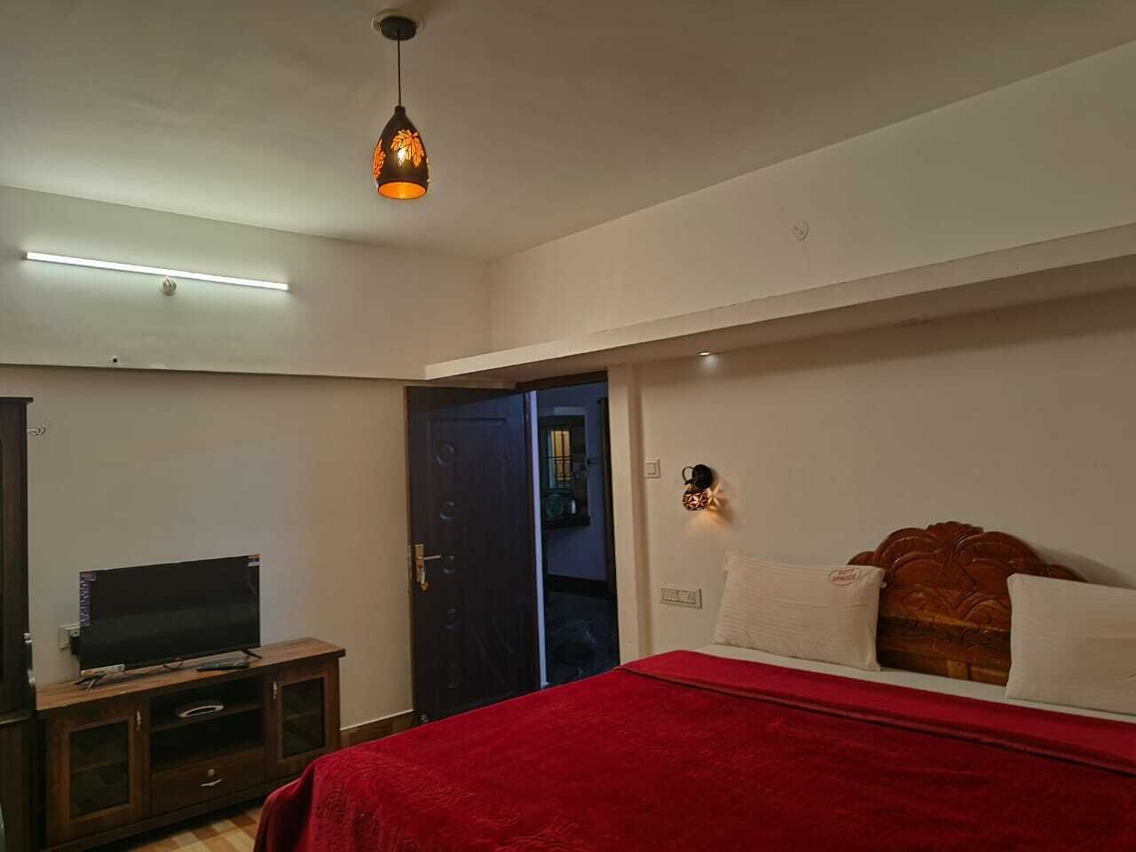 Um quarto com cama, televisão sobre suporte de madeira, manta vermelha, luminária de parede decorativa e porta que dá para outro cômodo.