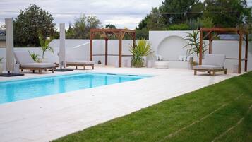 Piscina com espreguiçadeiras e pérgola, cercada por um muro branco e vegetação.