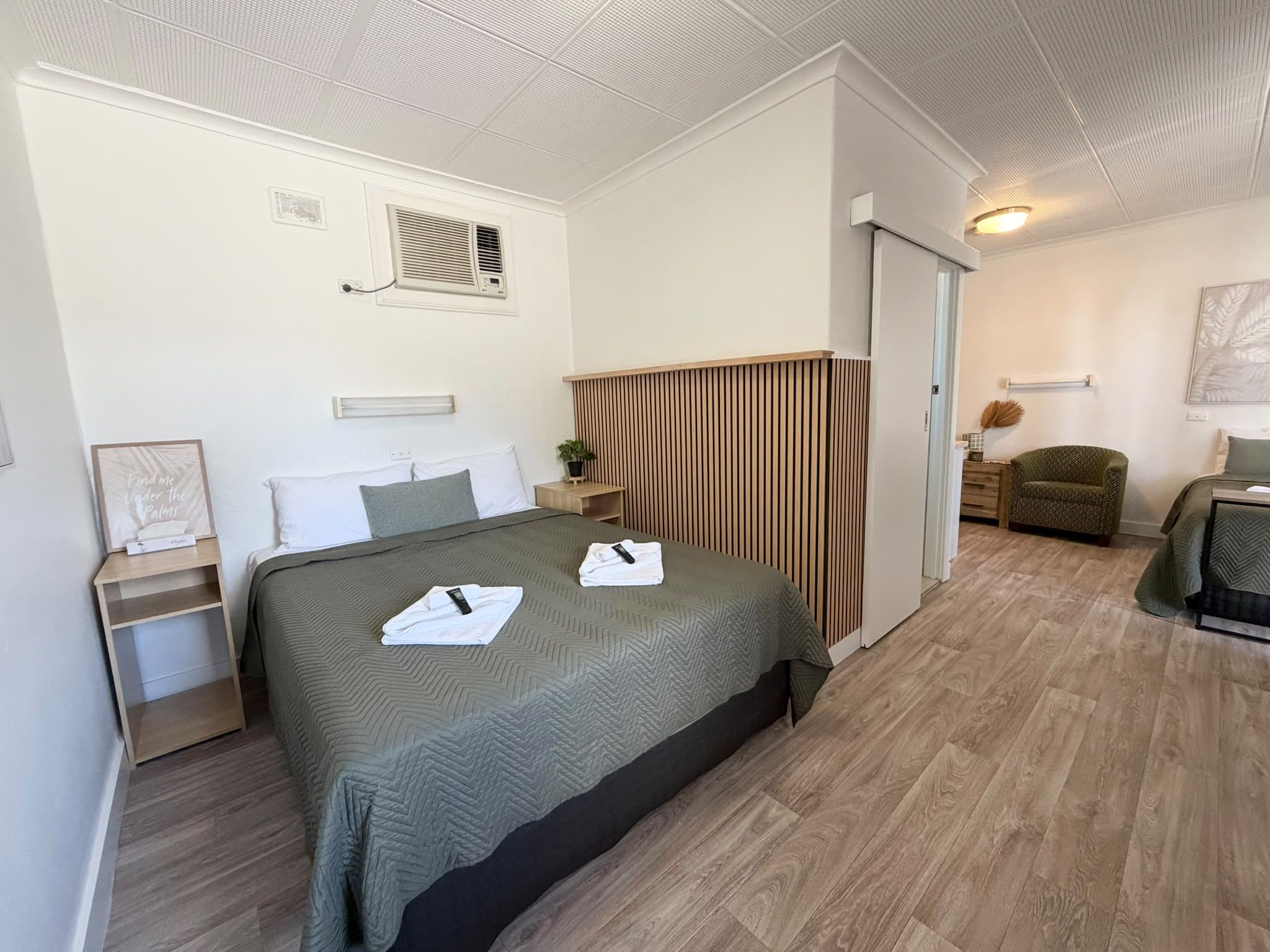 Quarto de hotel com cama, mesas de cabeceira, uma planta, uma cadeira e uma porta que dá para outro quarto.