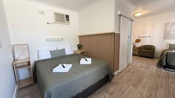 Quarto de hotel com cama, mesas de cabeceira, uma planta, uma cadeira e uma porta que dá para outro quarto.