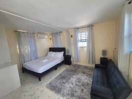 Um quarto com cama, sofá, mesa de cabeceira, ar condicionado e janela com cortinas.