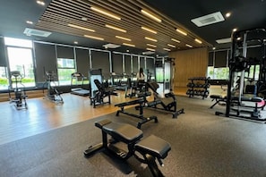 Academia com diversos equipamentos de exercícios, janelas amplas e teto de madeira.