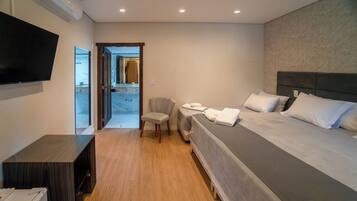 Quarto de hotel com cama, televisão, poltrona e escrivaninha.
