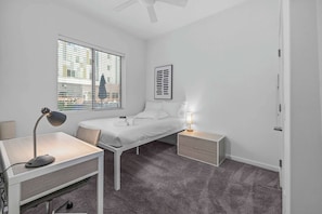 Quarto com cama, mesa de cabeceira, escrivaninha, cadeira, janela e ventoinha no teto.