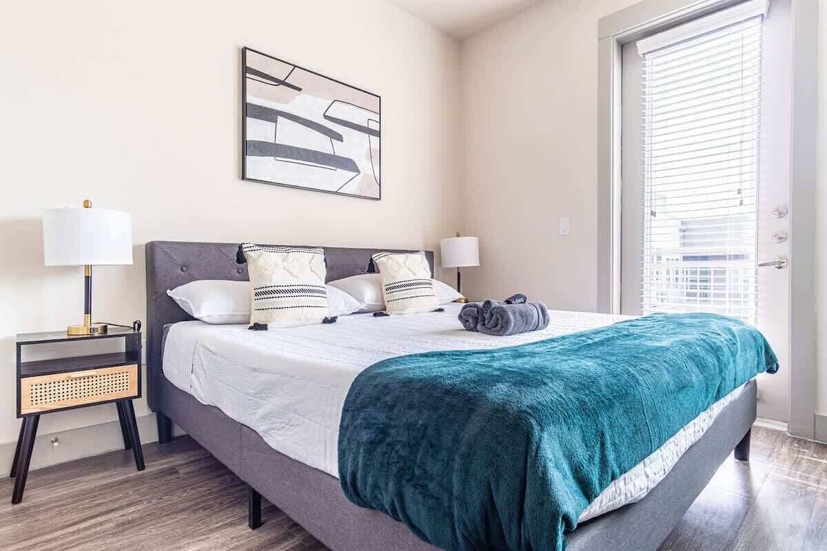 Quarto com cama, duas mesas de cabeceira, janela com persianas e uma obra de arte abstrata na parede.