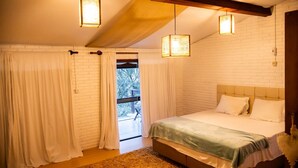 Quarto com cama, cortinas brancas, vista por uma porta de vidro e luminárias pendentes.