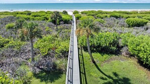 Um calçadão leva a uma praia com vegetação densa e uma vista clara do oceano.