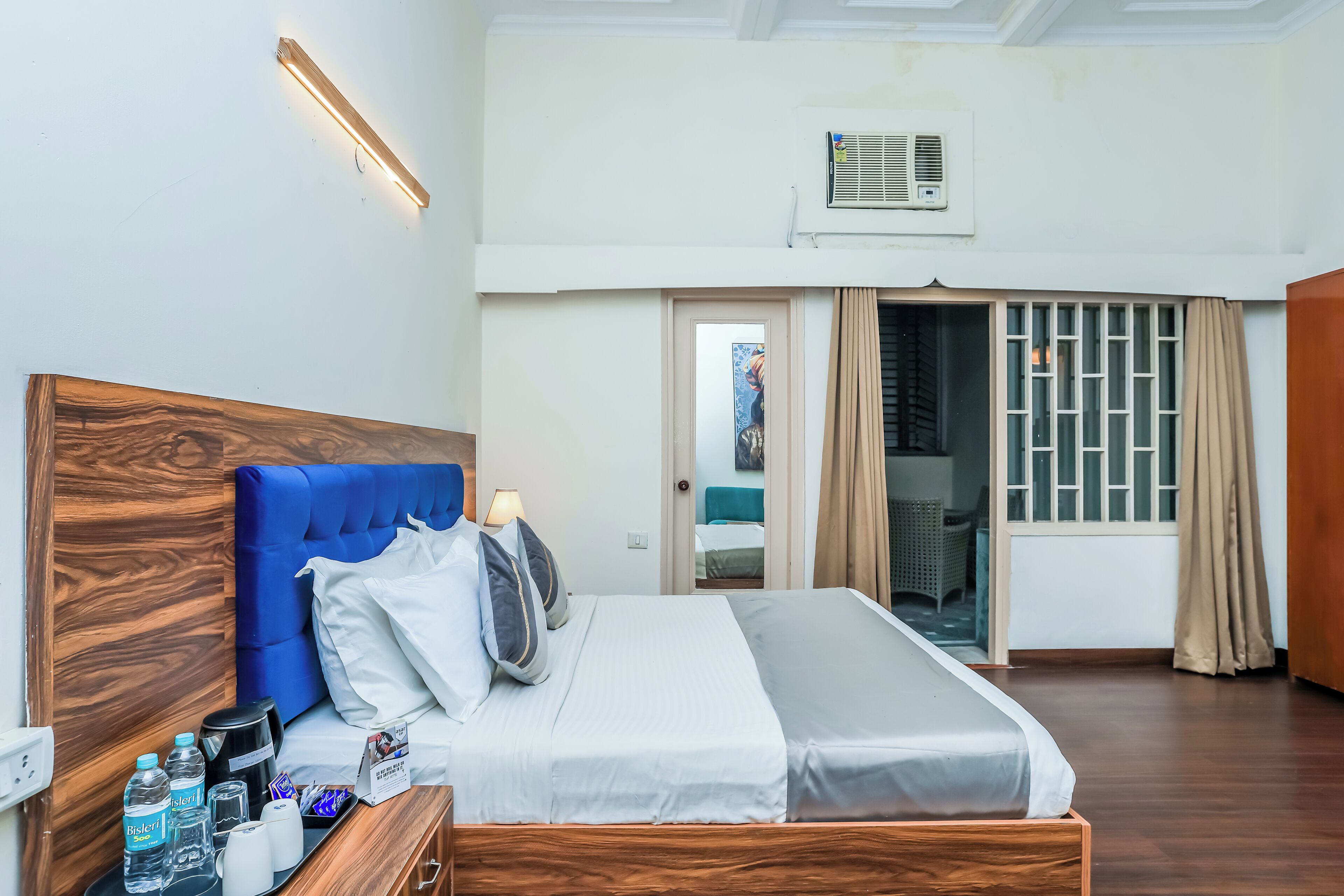 Quarto de hotel com cabeceira de madeira, cama com roupa de cama branca e cinza, mesa de cabeceira com garrafas de água, cafeteira e abajur.