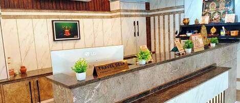Balcão de recepção com placa indicando "TRAVEL DESK", uma imagem emoldurada de uma divindade e um pequeno santuário com oferendas.