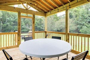 Um deck coberto ao ar livre com mesa e cadeiras redondas, cercado por grades de madeira e vista para uma área florestal.