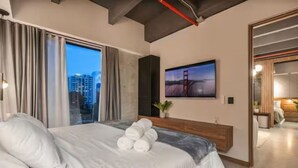 Quarto de hotel moderno com uma cama grande, televisão de tela plana, vista para a cidade e cabeceira de madeira.