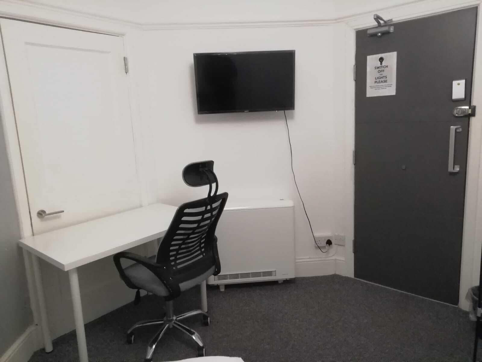 Um pequeno escritório com uma mesa branca, uma cadeira de escritório preta, uma televisão de tela plana fixada na parede e uma porta com uma placa indicando para desligar as luzes.
