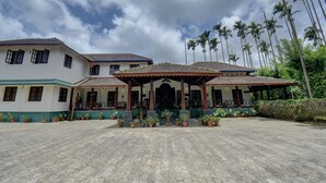 Um edifício em estilo colonial com um pavilhão central, rodeado por um pátio pavimentado e palmeiras.