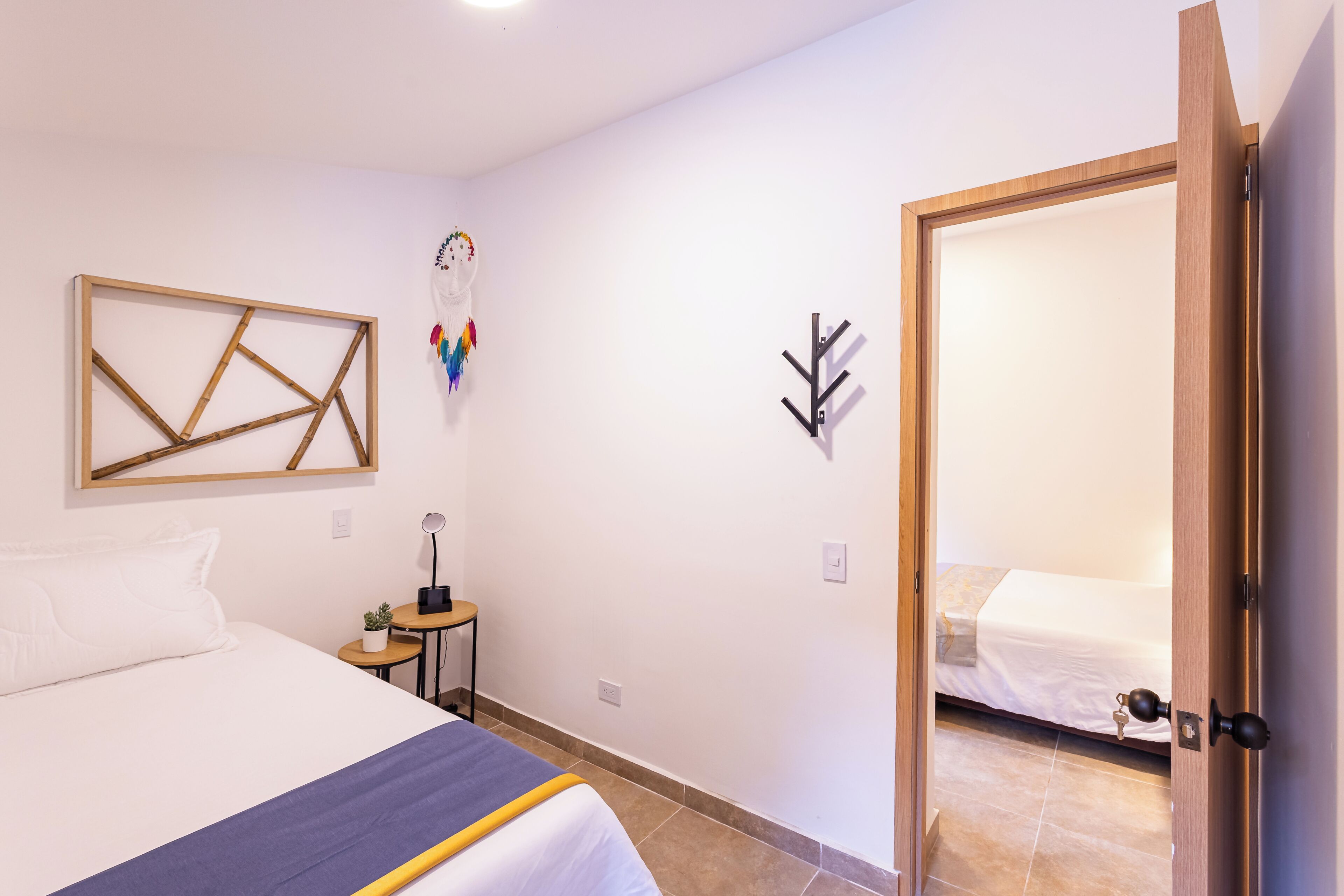 Quarto de hotel com cama, mesa de cabeceira, luminária, arte na parede e porta que dá para outro cômodo.