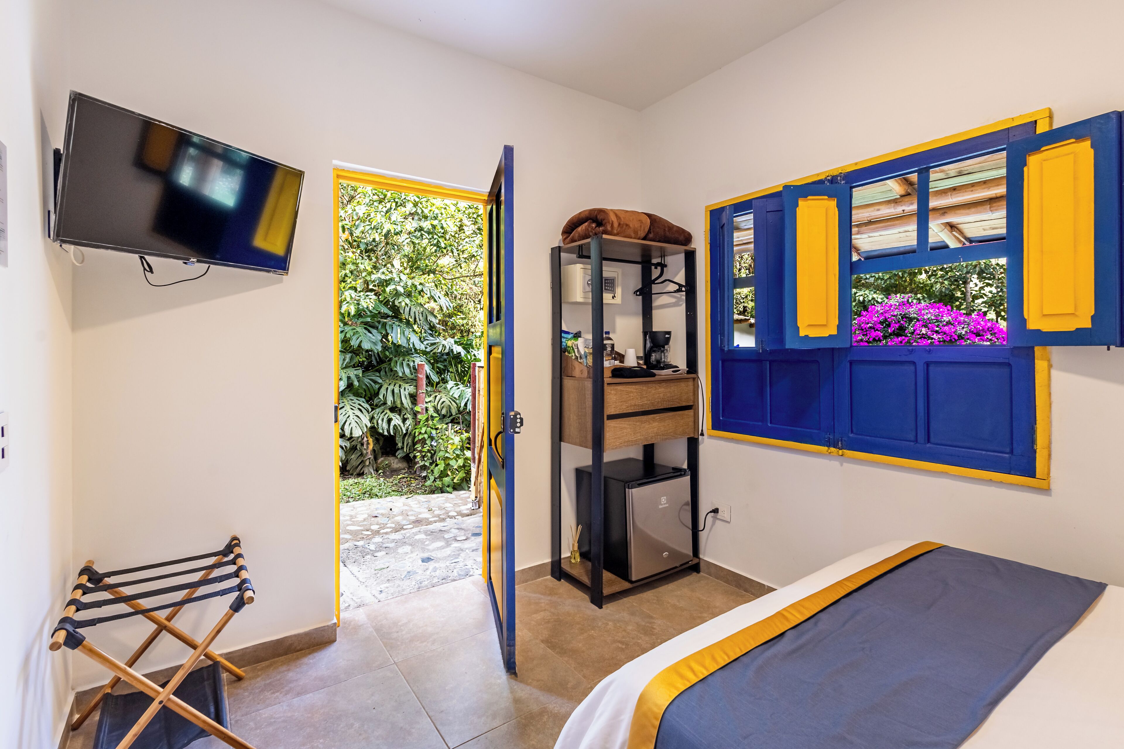 Quarto com cama, televisão na parede, prateleira com objetos e janela com persianas azuis.