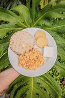 Um prato com alimentos, incluindo pão achatado, ovos mexidos e biscoitos, em frente a um fundo de folhas verdes grandes com gotas de água.