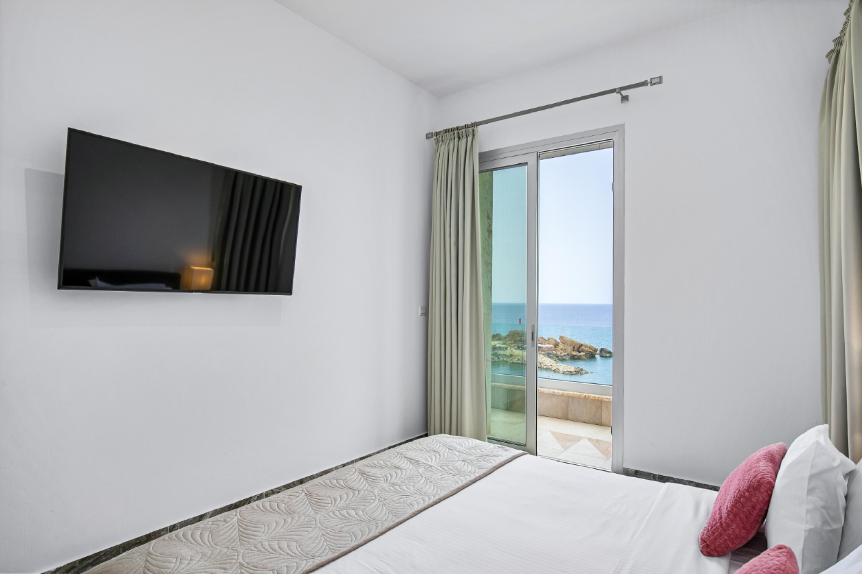 Quarto com cama, TV fixada na parede e porta de vidro deslizante para uma varanda com vista para o mar.