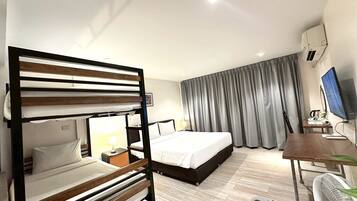 Quarto de hotel com uma cama beliche, uma cama de solteiro, uma escrivaninha com um computador, uma TV e uma janela grande com cortinas.
