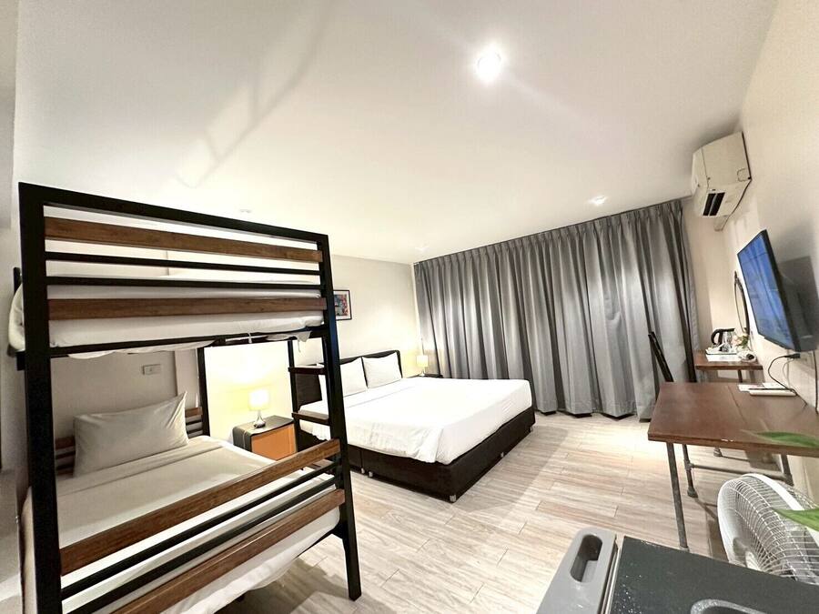 Quarto de hotel com uma cama beliche, uma cama de solteiro, uma escrivaninha com um computador, uma TV e uma janela grande com cortinas.