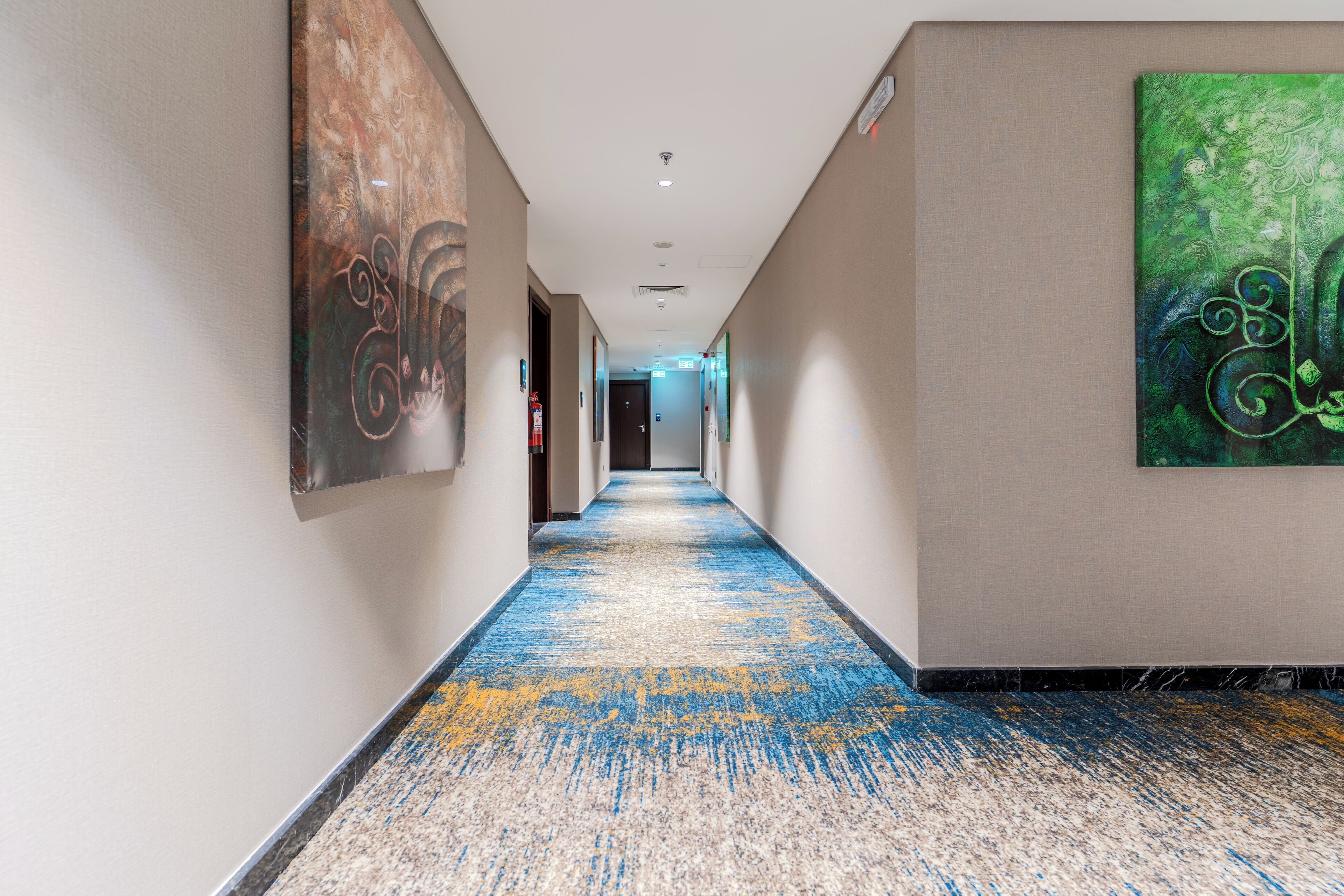 Corredor de hotel com carpete estampado, obras de arte nas paredes e um extintor de incêndio.