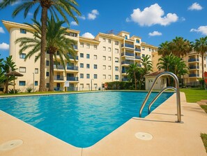 Piscina com escada de metal, rodeada por palmeiras e um edifício com várias andares e sacadas.