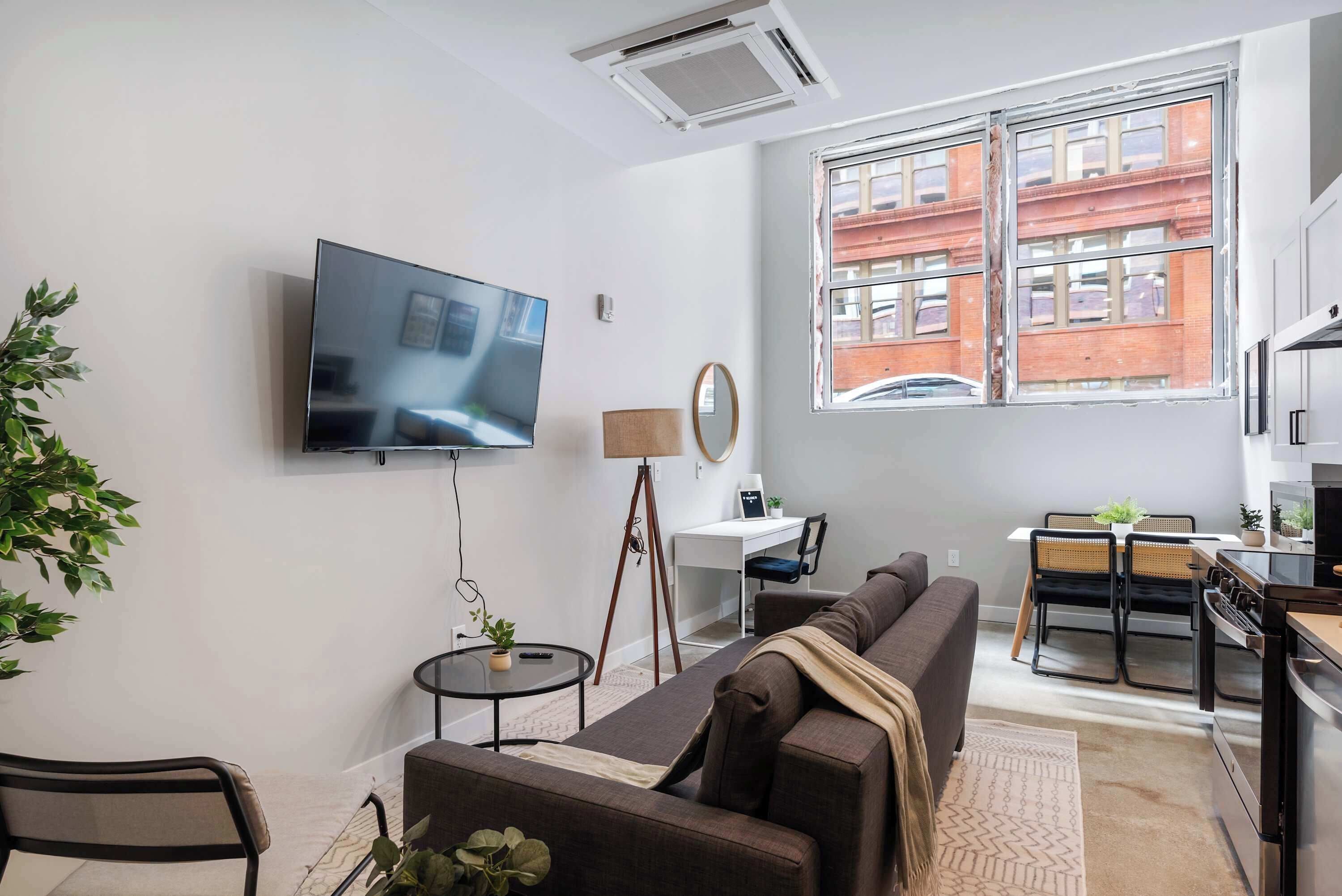 Uma sala de estar moderna com televisão de tela plana, um sofá, uma área de jantar com mesa e cadeiras, e uma cozinha com fogão.