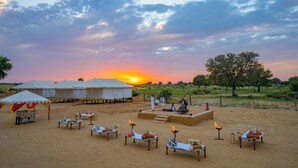 Acampamento no deserto com tendas, cadeiras de descanso e um palco central com pessoas.
