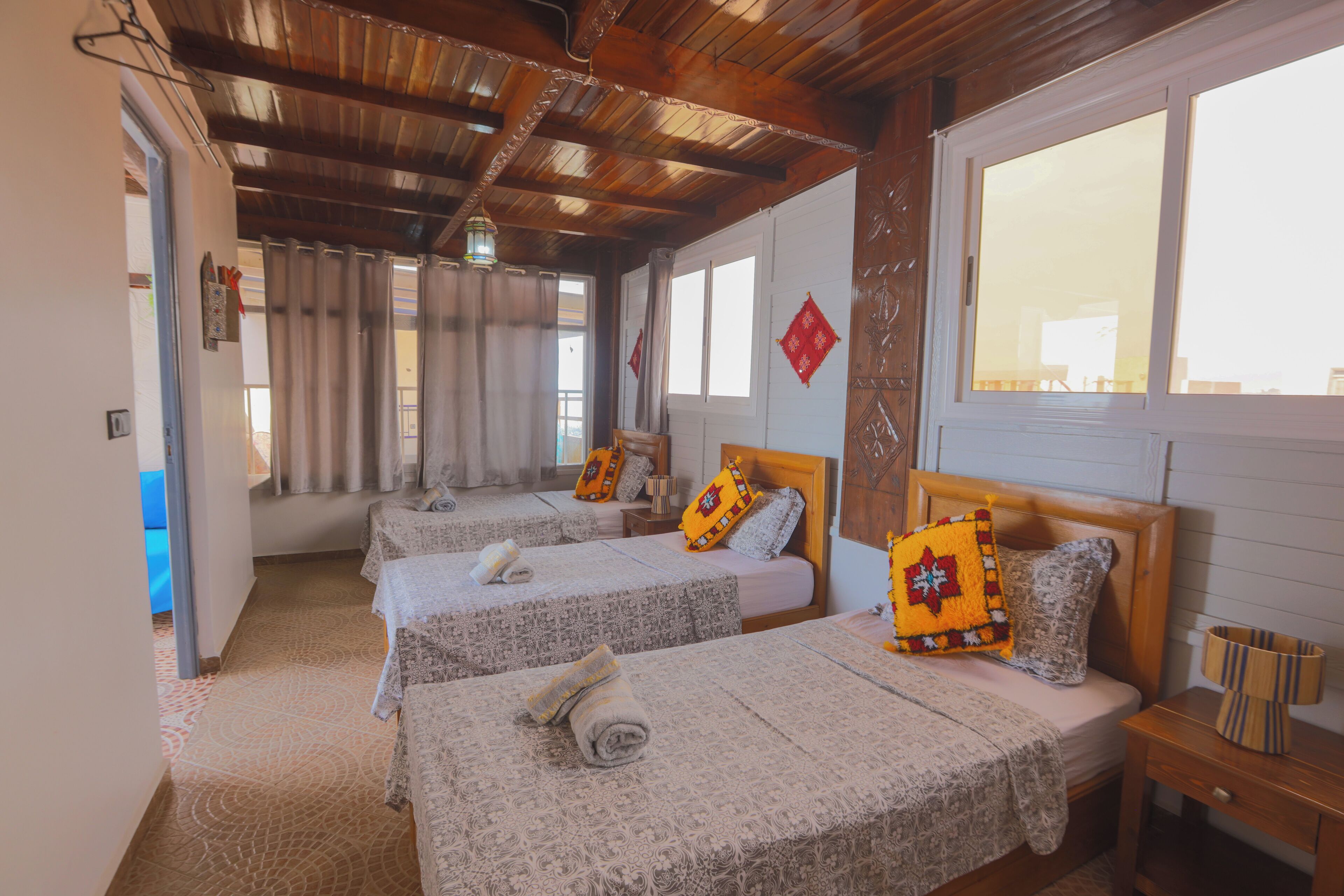 Quarto de hotel com três camas, cada uma com colcha e almofadas estampadas, cabeceira de madeira e mesa de cabeceira com luminária.